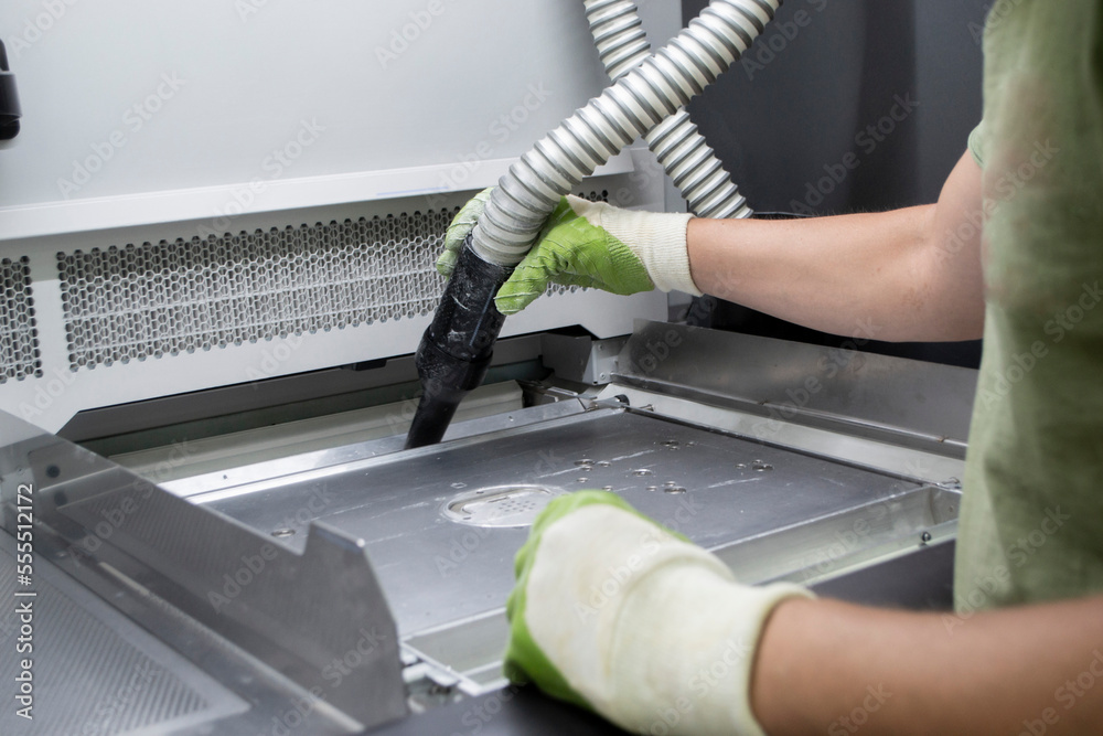 A male worker cleans the surface of an industrial 3D printer from white powder with a vacuum ...