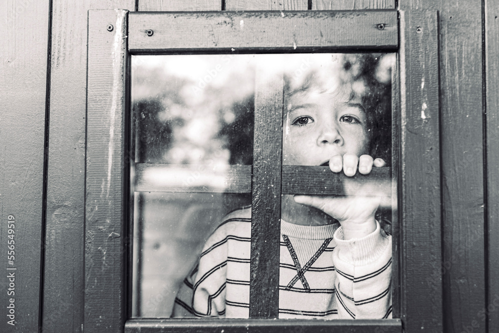 A beautiful little Caucasian boy locked in a wooden hut looks out the ...