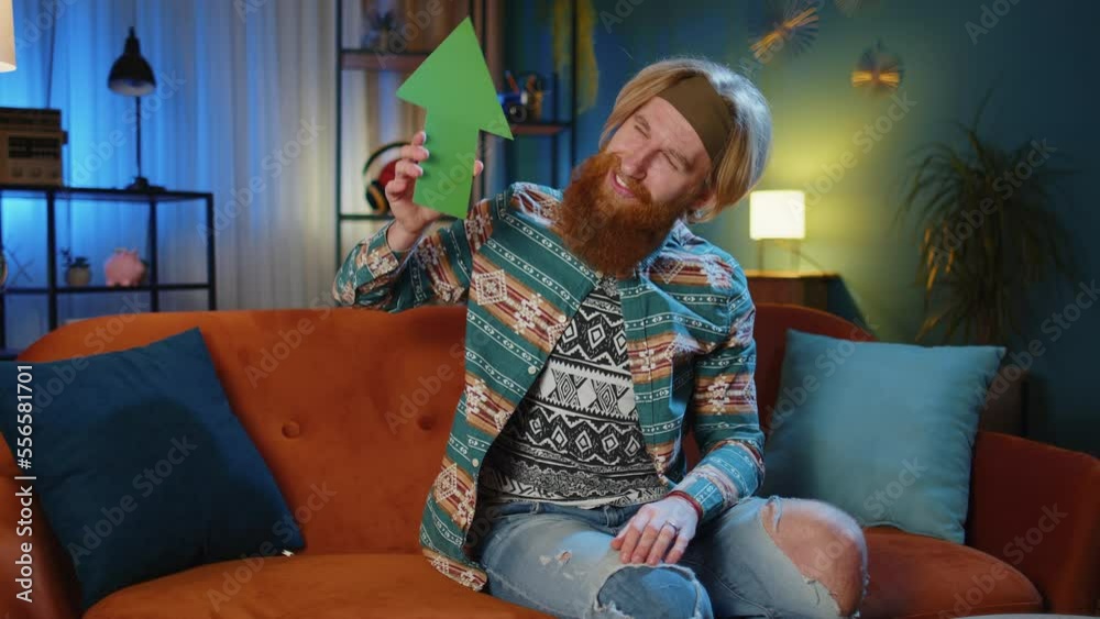 Happy young hippie man at home couch winner holding arrow sign pointing ...