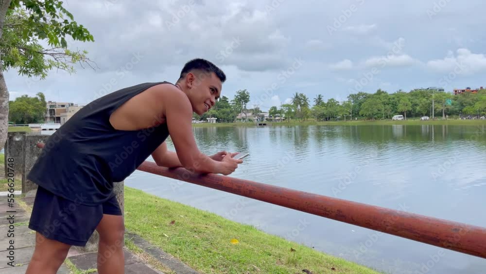 Filipino young man leaning on railing by lake using smart phone. Asian ...