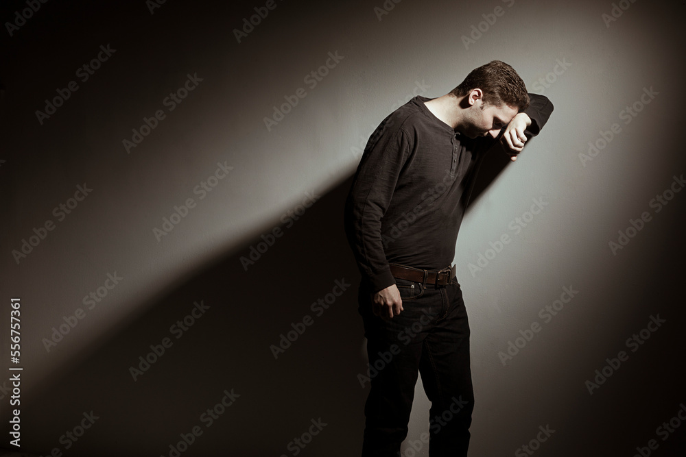 A young man stands leaning against a wall with his head on his arm ...