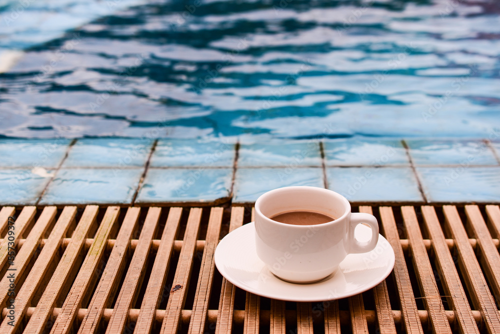 White cup of coffee by the edge of the pool, blurred blue water ...