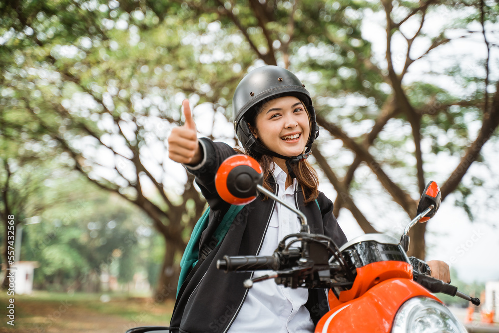 Student girl with thumbs up riding motorbike wearing helmet and jacket ...