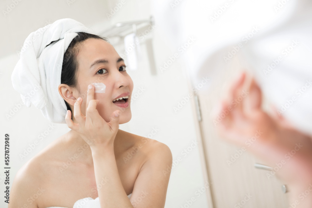 Happy young Asian woman applying face lotions while wearing a towel and touching her face in ...