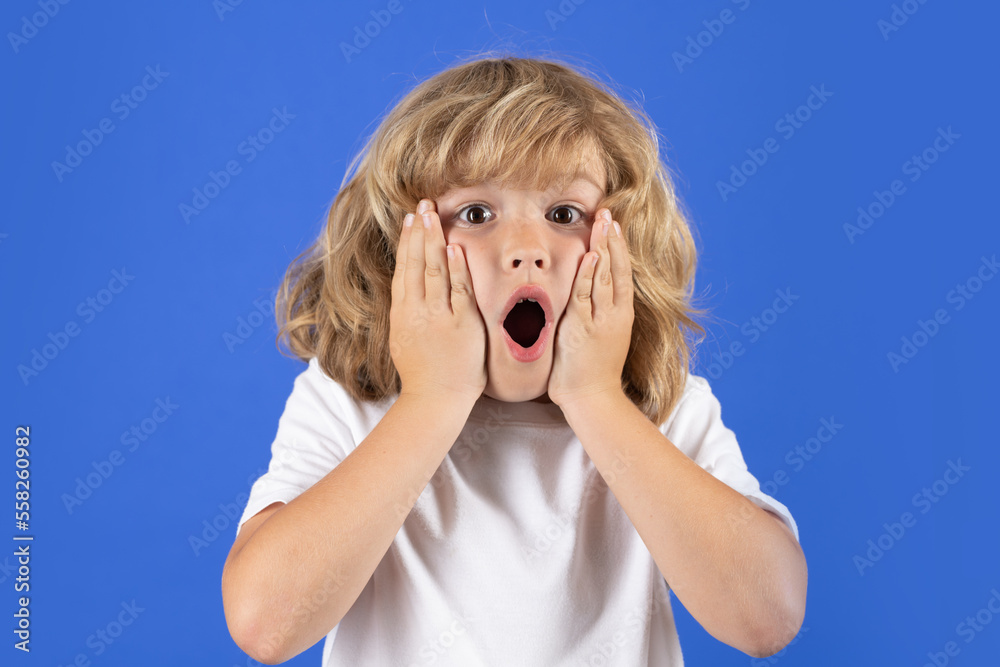 Shocked kid boy keeping hands near cheek with open mouth on blue ...