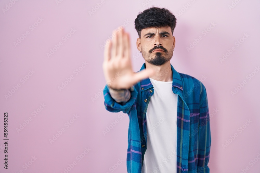 Young hispanic man with beard standing over pink background doing stop ...