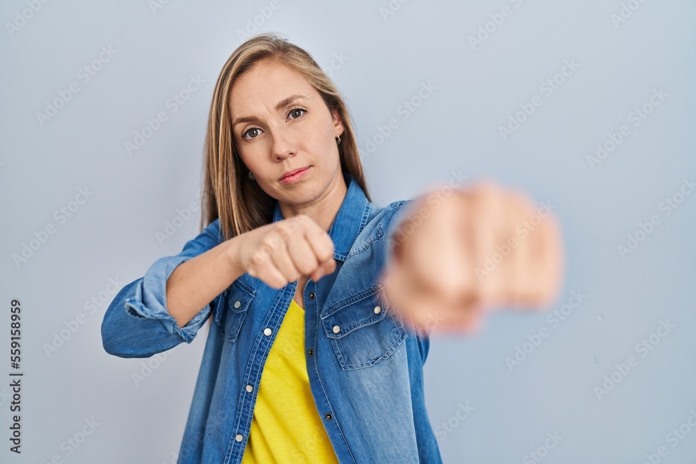 Young blonde woman standing over blue background punching fist to fight ...