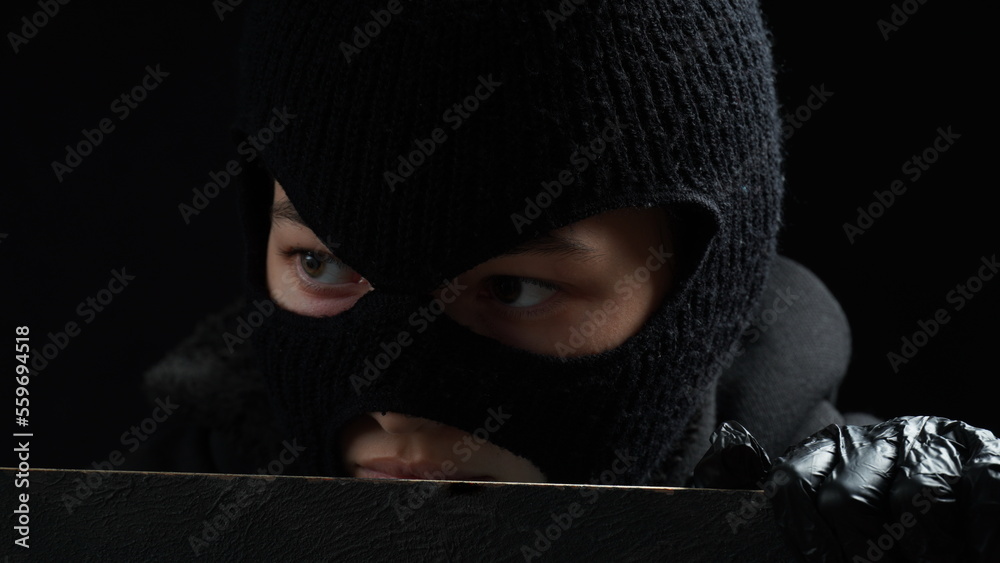 Masked female burglar in black gloves looks over a fence. Crime concept ...
