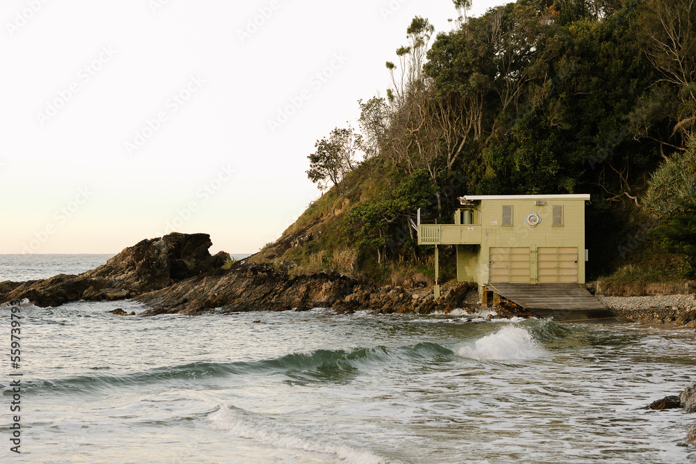 Small green boat shed on the beach with small waves and a hill behind ...