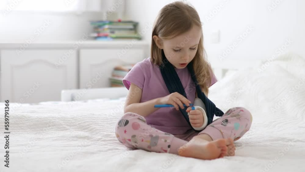 Adorable preschooler girl with a broken arm at home on the bed draws ...