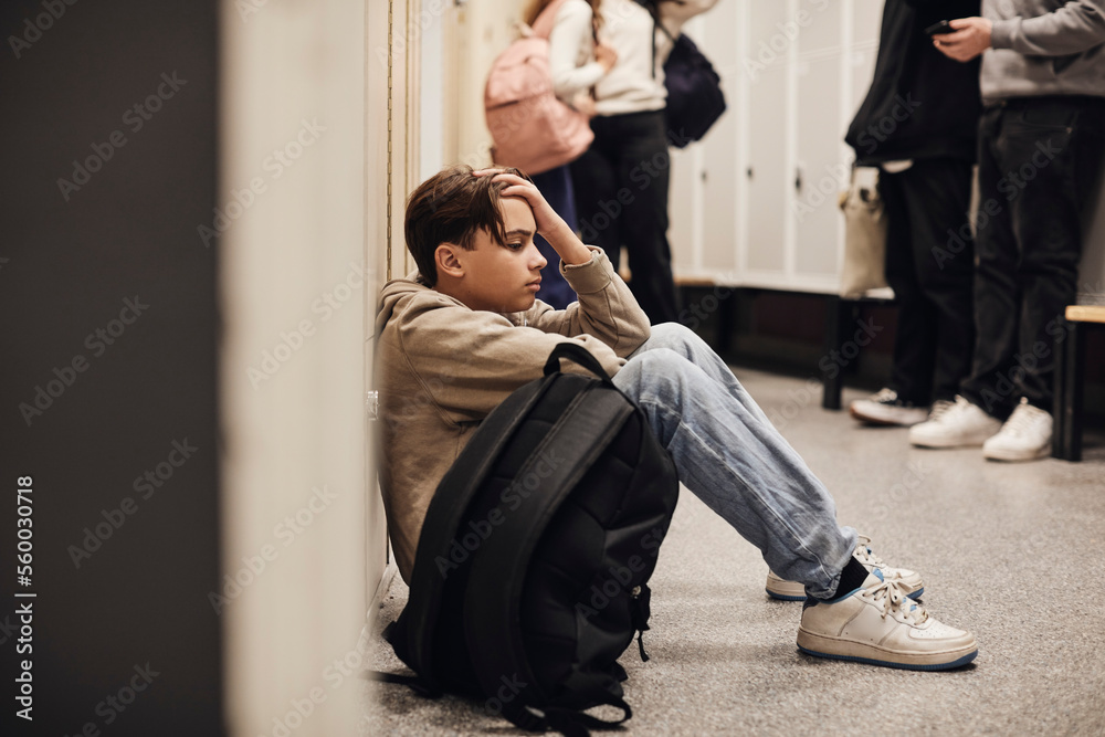 Side view of sad teenage boy sitting with backpack in school corridor ...
