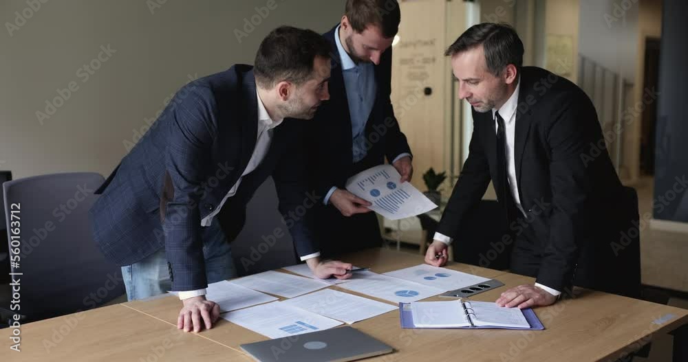 Group of businessmen in suits work together in modern office boardroom, analyzing stats shown on ...