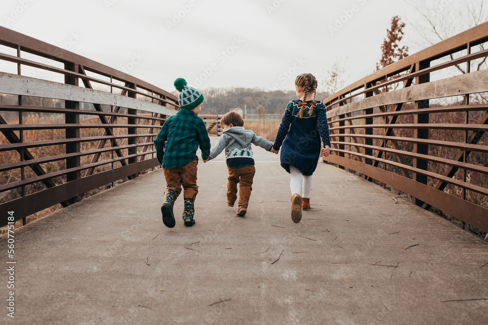 Three siblings holding hands running on bridge away from camera Stock ...
