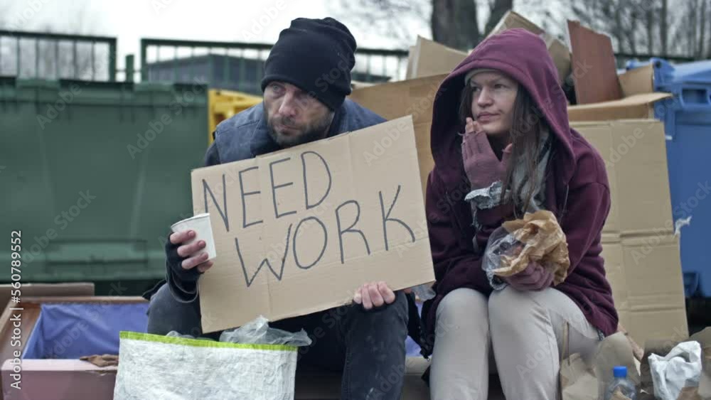 Two sad homeless men sit by the rubbish bins. Man and a woman are poor ...