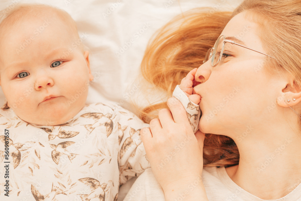 Cheerful young mother kiss fingers on palm of little baby lying on bed ...