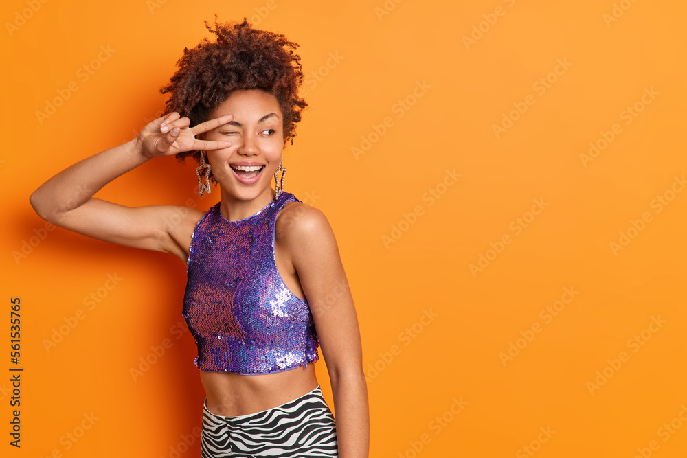 Horizontal shot of positive curly haired woman shows peace sign smiles ...