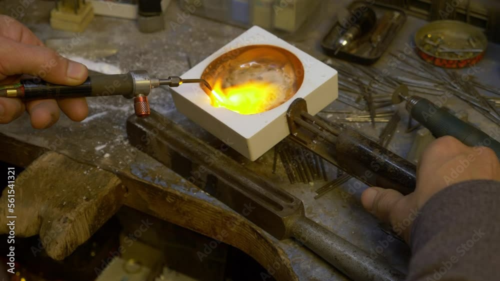 Goldsmith heating a bar of casted raw gold in a pan using a burner ...