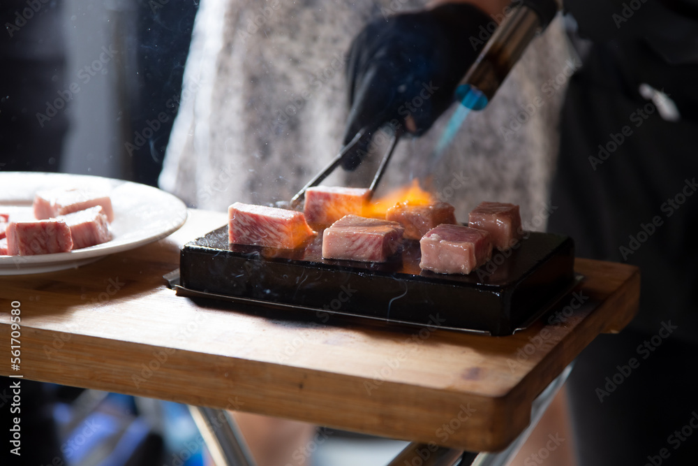 A view of a cook using a blow torch on cubes of A5 wagyu beef on a hot ...