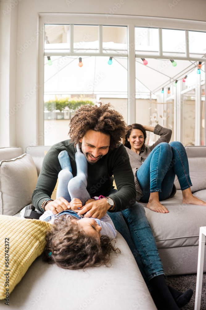 Happy father tickling daughter on sofa with mother sitting in ...
