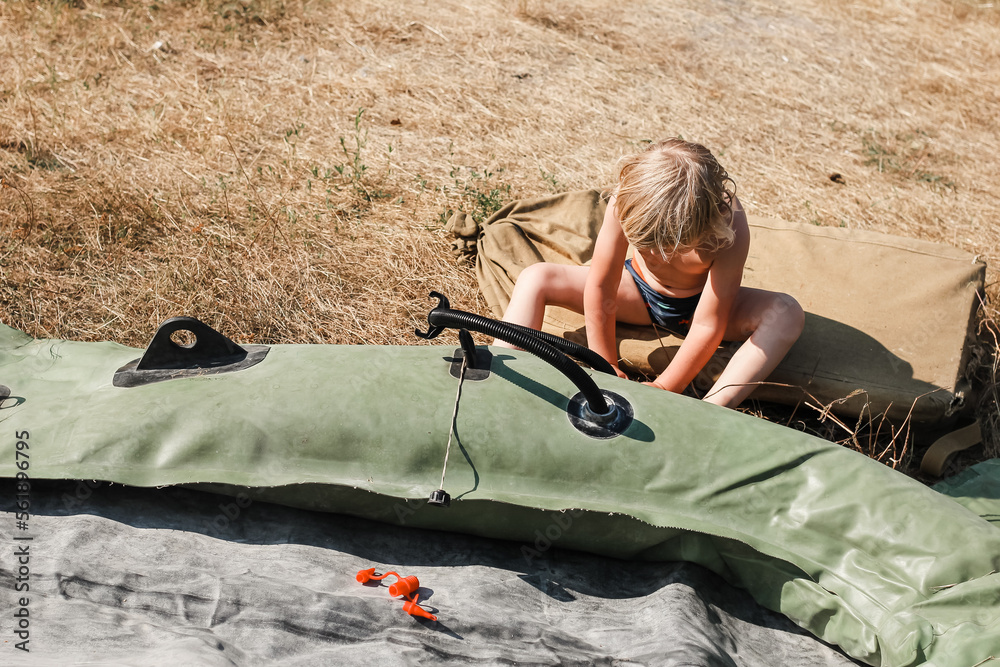 A little blonde boy inflating a rubber boat with a pump during family ...