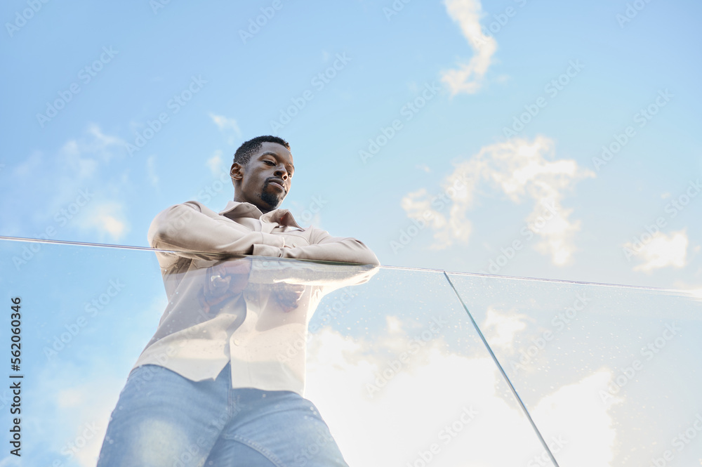 Handsome African young man leaning on a glass railing with a thoughtful ...