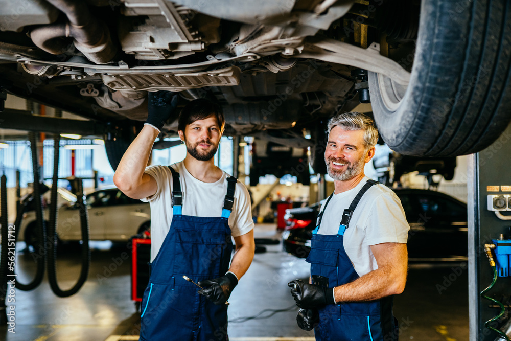 Two smiling mechanics, young and older men , stand under a car seen on ...