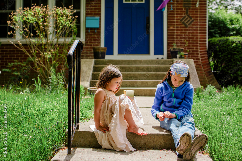 Two little girls sit on front stoop in sunshine talking to each other ...