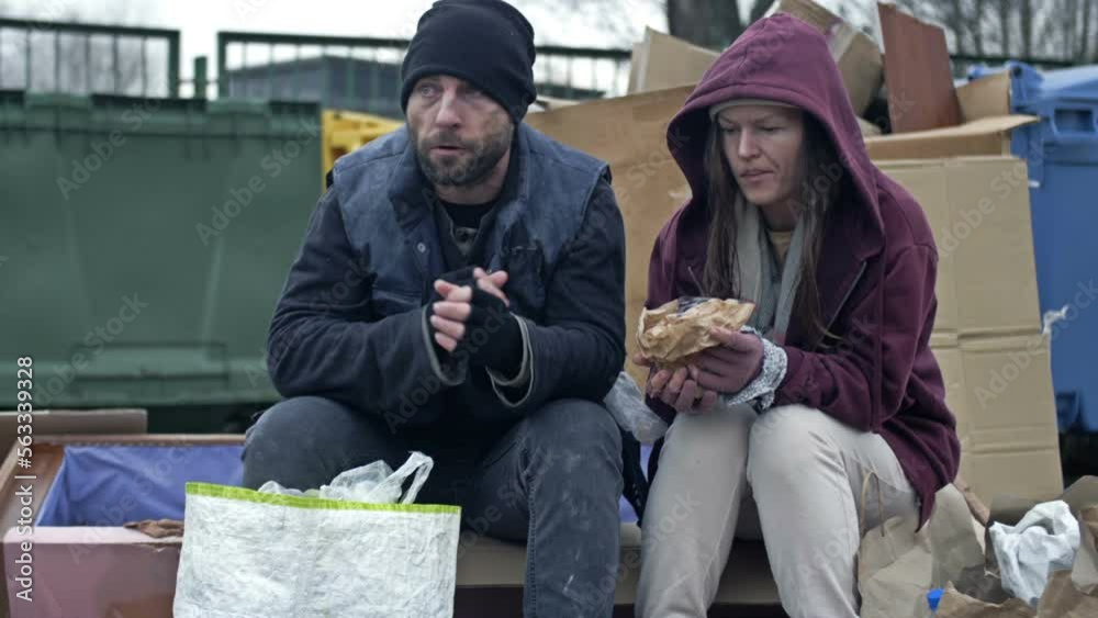 Two dirty and poorly dressed homeless people, a man and a woman, sit by ...