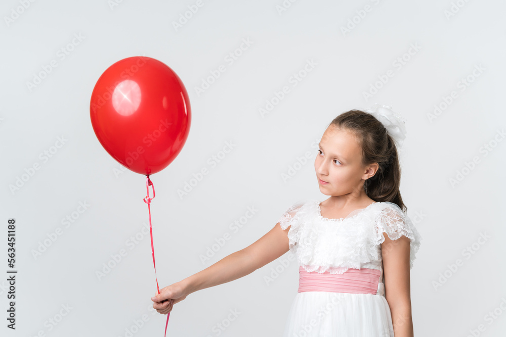 Pretty girl in white dress holding one red balloon in outstretched hand and looking thoughtfully ...