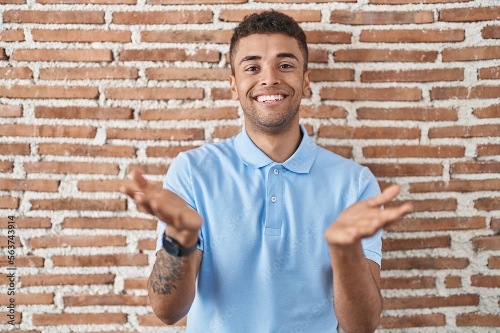Brazilian young man standing over brick wall smiling cheerful offering ...