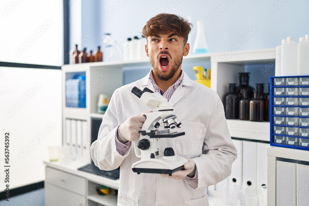 Arab man with beard working at scientist laboratory holding microscope angry and mad screaming ...