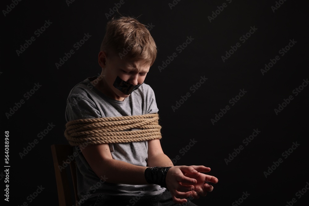 Little boy with taped mouth tied up and taken hostage against dark background. Space for text ...