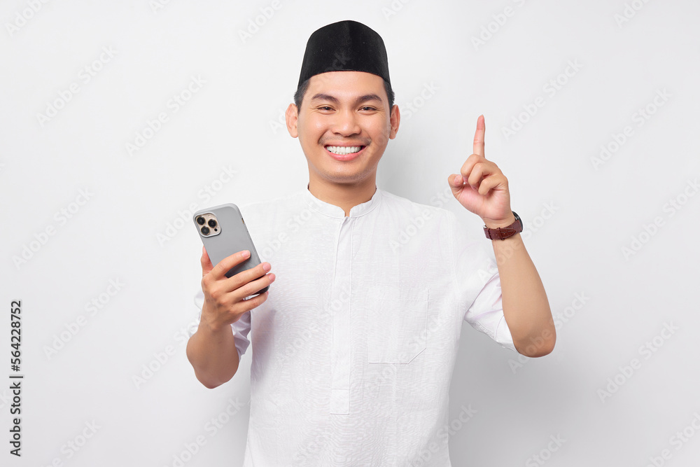 Smiling young Asian Muslim man holding mobile phone and pointing finger ...