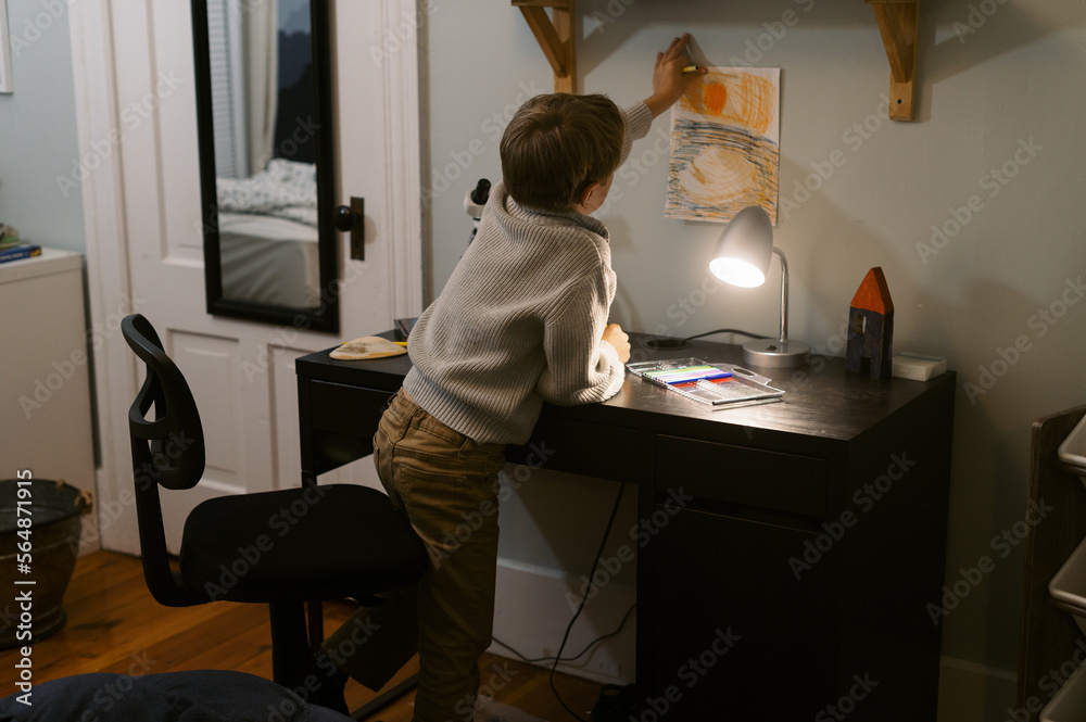 Little boy in his room at his desk holding his drawing to his wall ...