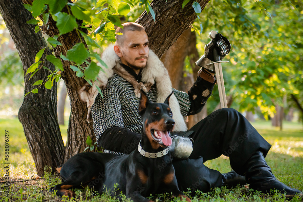 A young man in chain mail with a sword in the forest sits under a tree ...