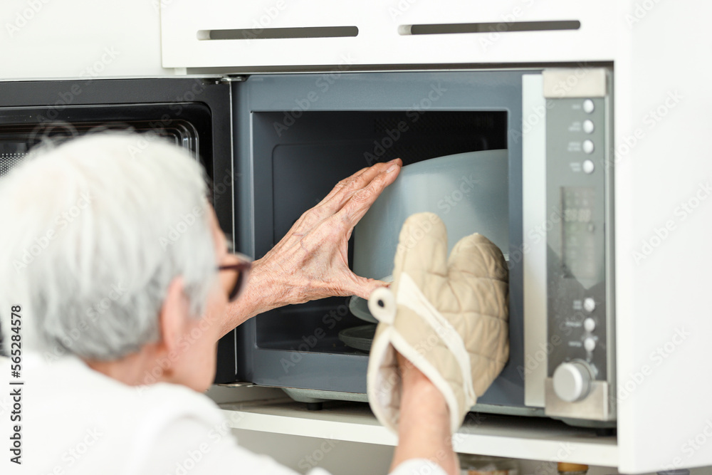 Asian senior woman wearing oven glove while taking food out of ...