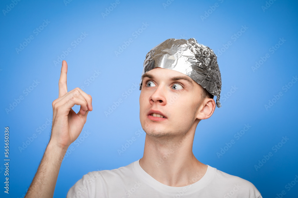 Paranoid young man wearing tin foil hat pointing up studio shot on blue ...