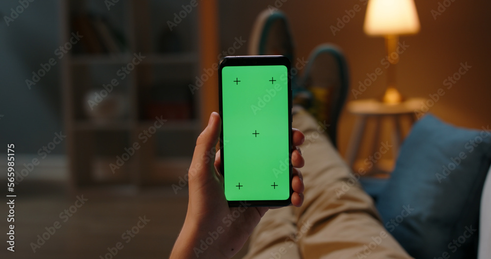 Man lying on couch using smartphone with chroma key green screen at night, scrolling through ...