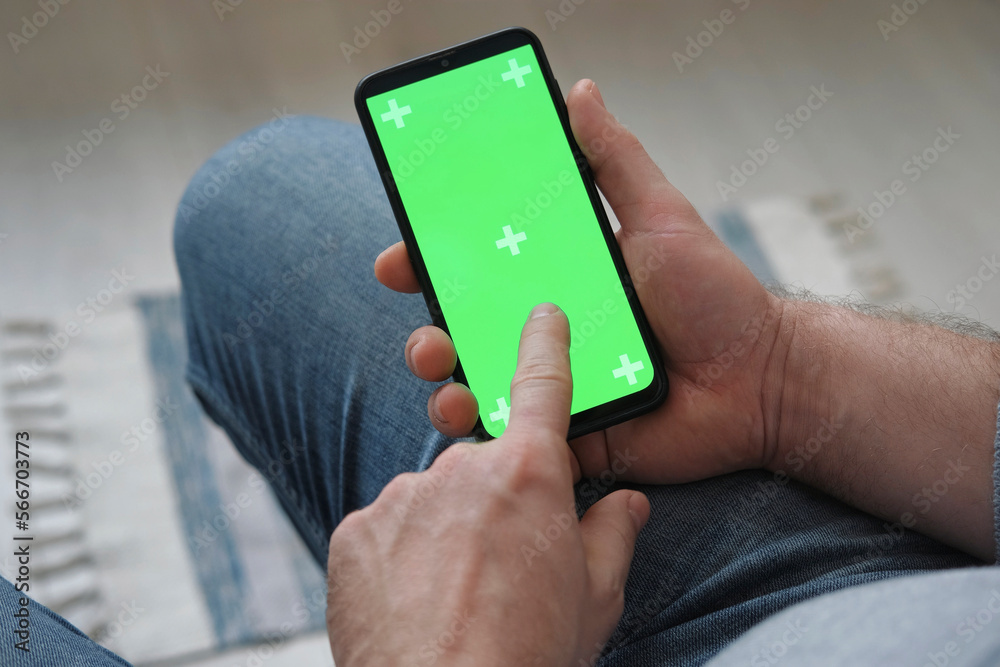 Young man sitting at home holding smartphone green mock-up screen in ...