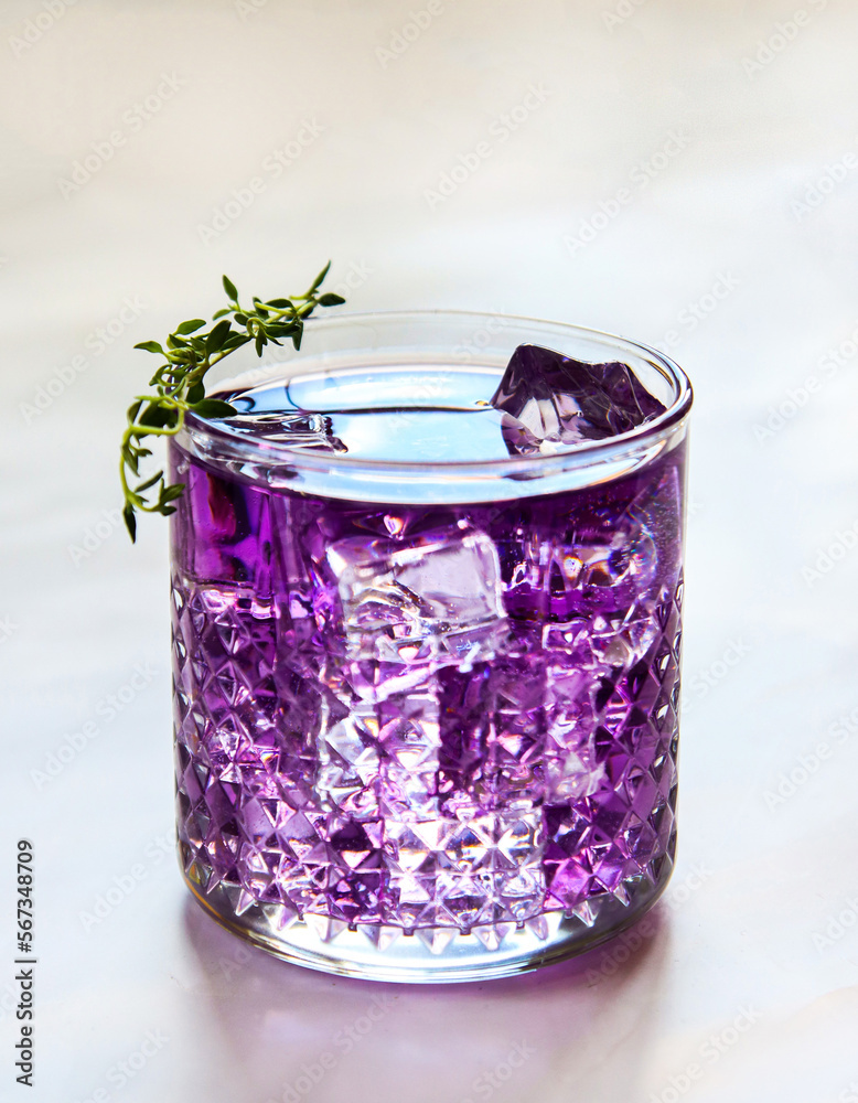 Purple drink in a transparent drinking glass with ice cubes and a thyme ...