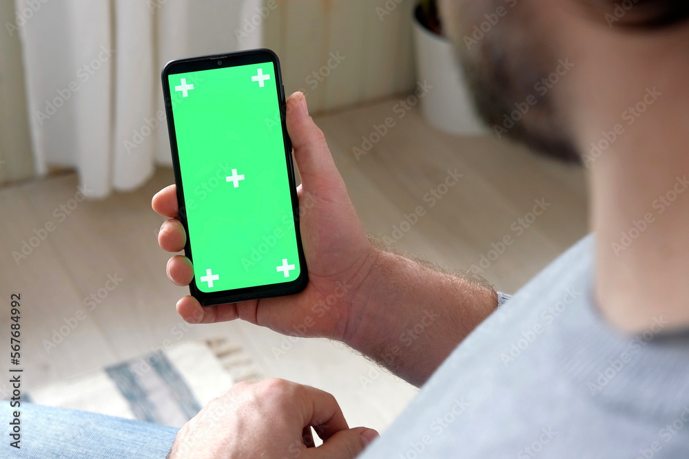 Young man sitting at home holding smartphone green mock-up screen in ...