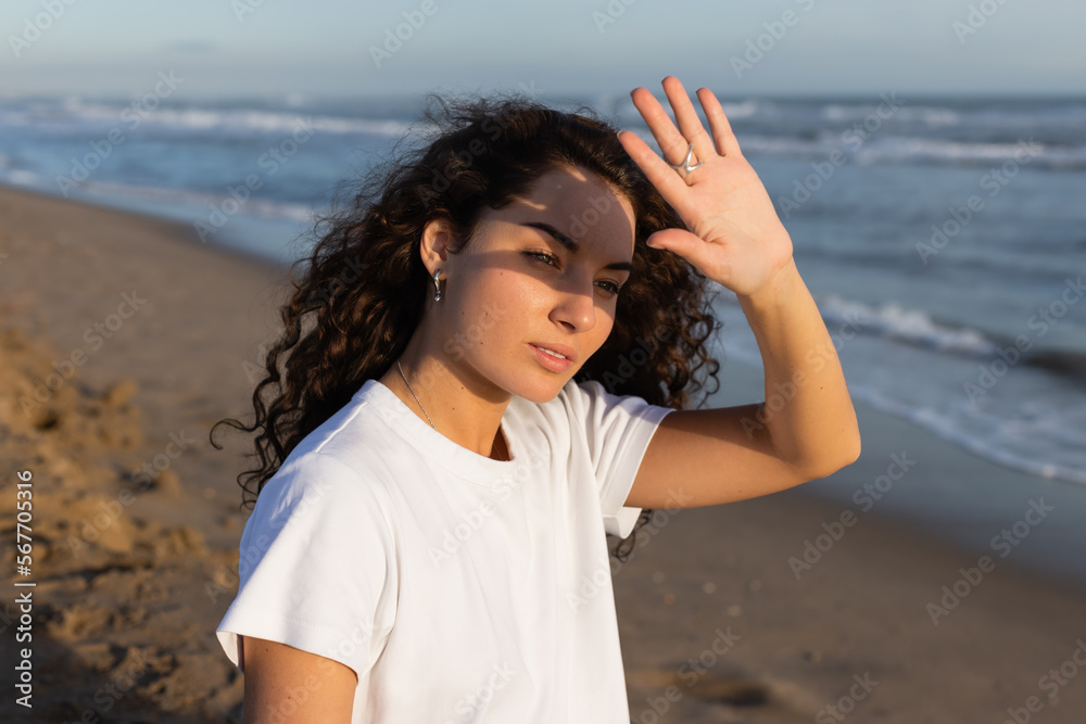 portrait of curly woman in white t-shirt hiding face from sun on beach ...