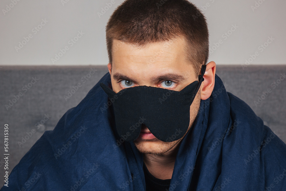 Embarrassed young man in a blanket at home wears a sleep mask while ...
