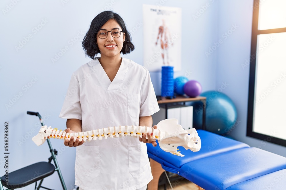 Young latin woman wearing physiotherapist uniform holding anatomical ...