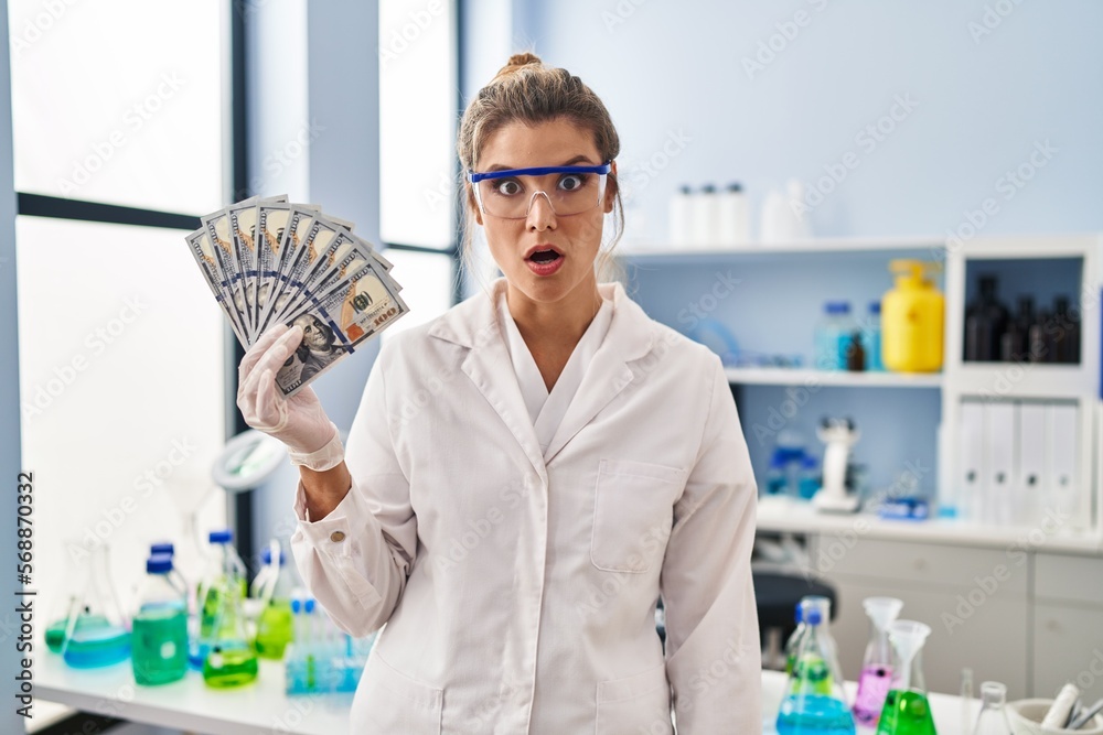 Young woman working at scientist laboratory holding money scared and ...