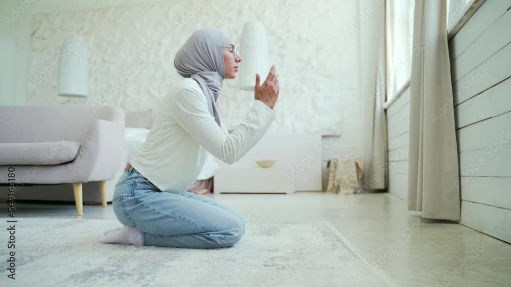A young Muslim woman in a hijab prays at home. Alone in a bright living ...