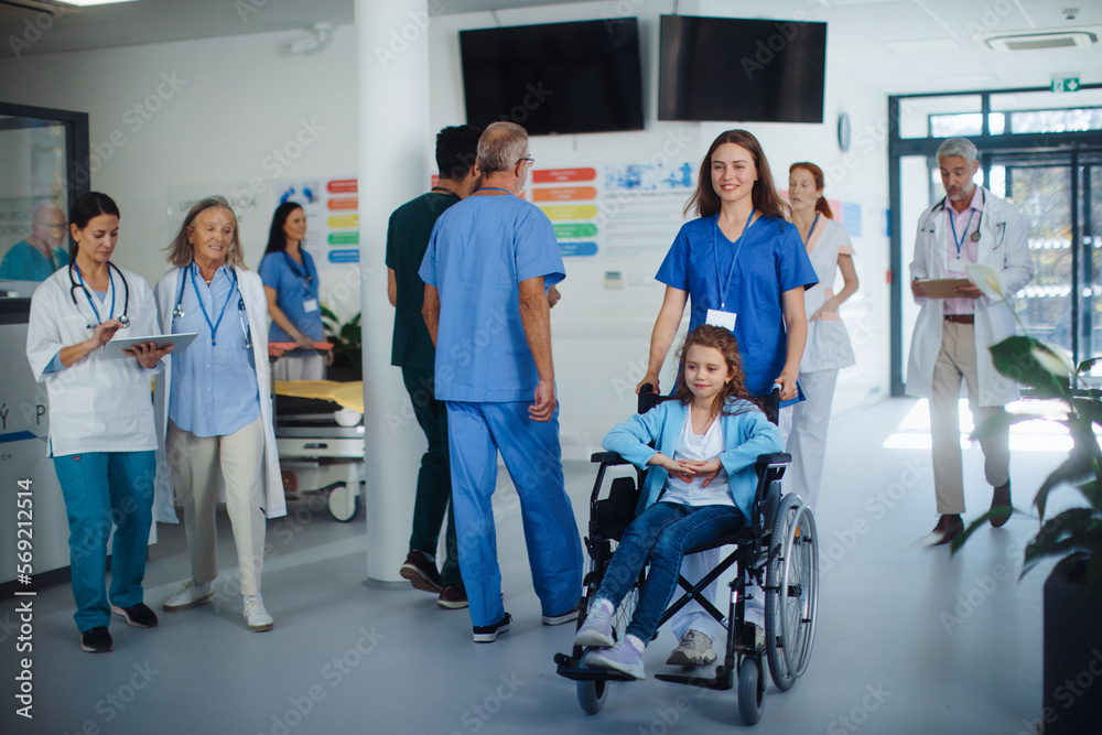 Young nurse pushing little girl on wheelchair at hospital corridor ...