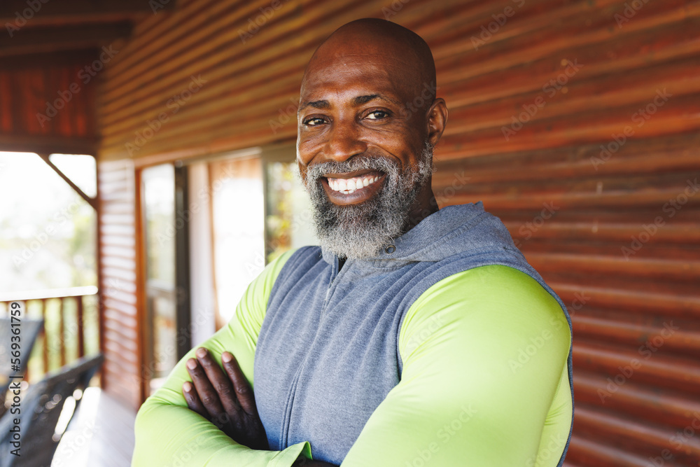 Smiling bald african american senior man with arms crossed standing against wall in log cabin ...