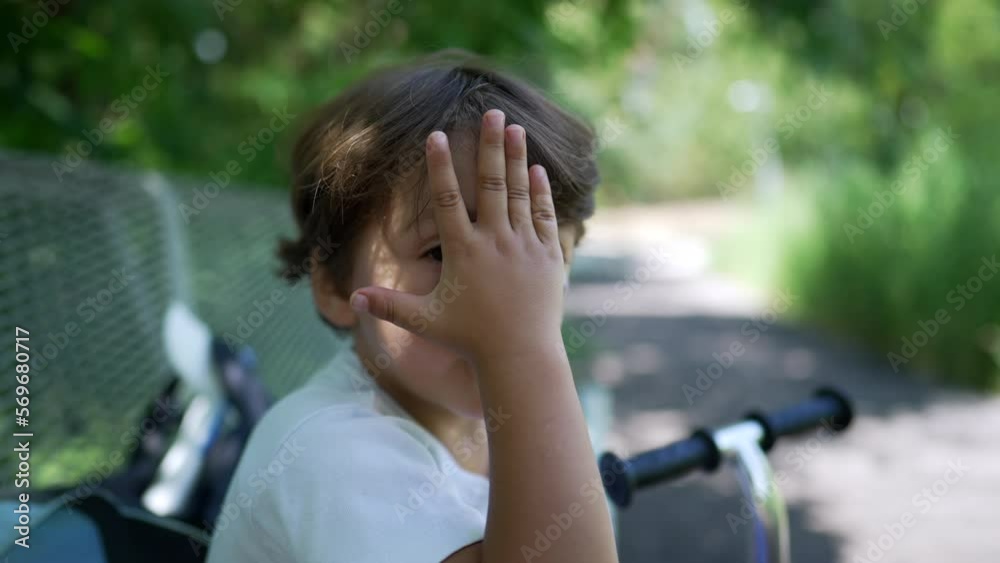 Little boy rubbing nose with hand seated at park bench. Kid touching ...