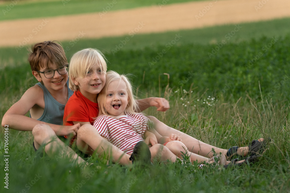 Three children sit on green grass and smile. Siblings outside. Two ...